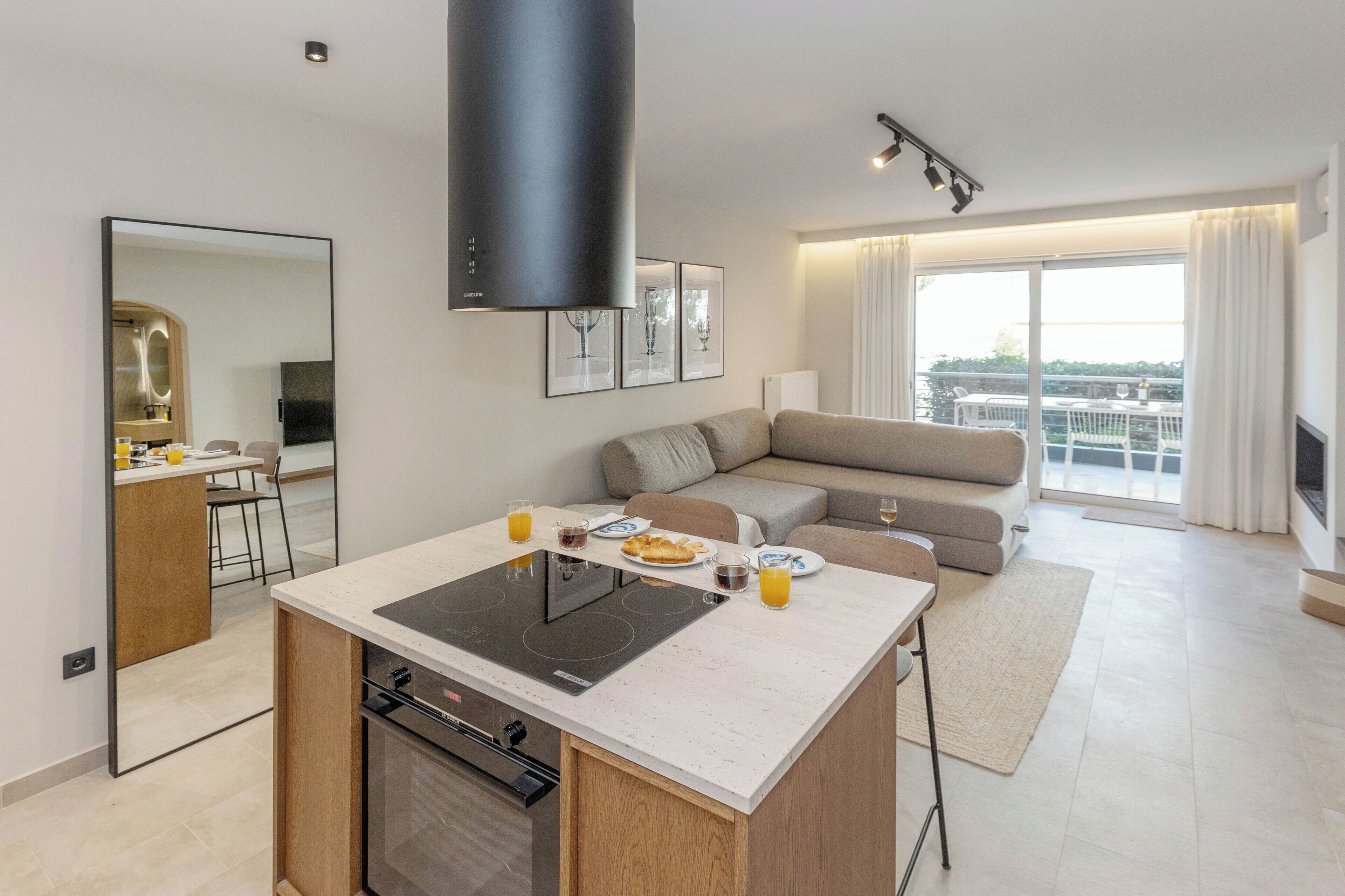 Uma cozinha moderna com uma ilha central, área de refeições com mesa e cadeiras, sala de estar com sofá e uma janela grande com cortinas.