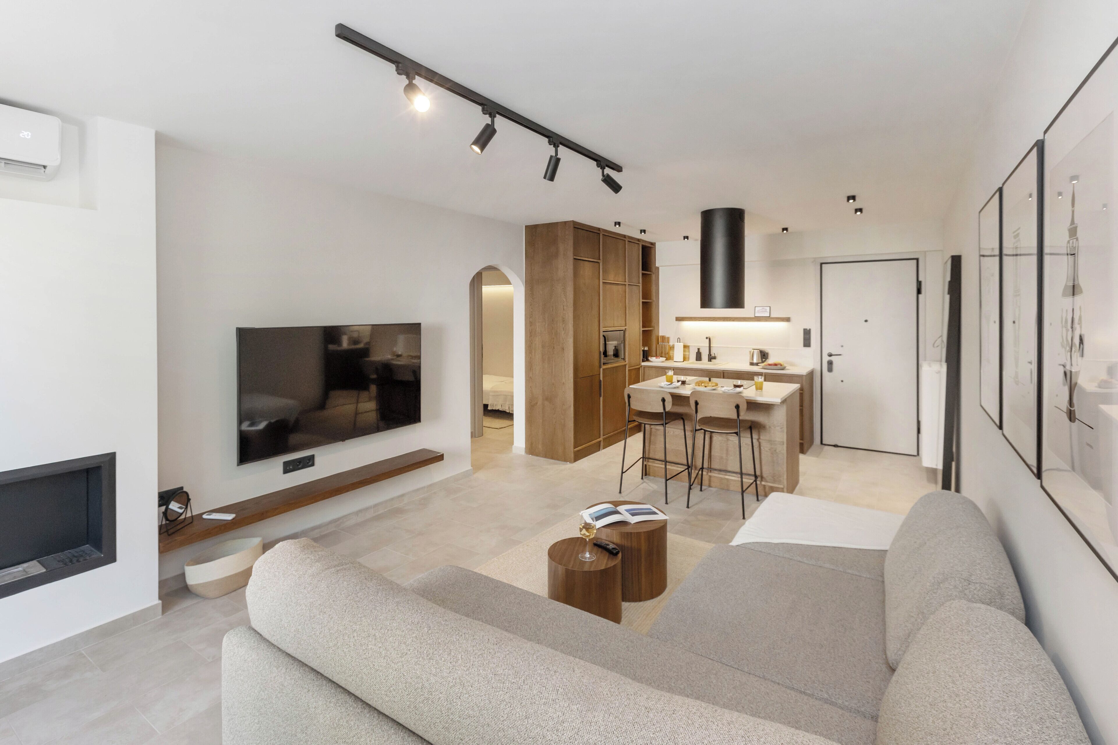 Uma sala de estar moderna com uma grande TV de tela plana, um sofá e uma mesa de centro. Há uma cozinha com um balcão e banquetas, e uma mesa de jantar com cadeiras.