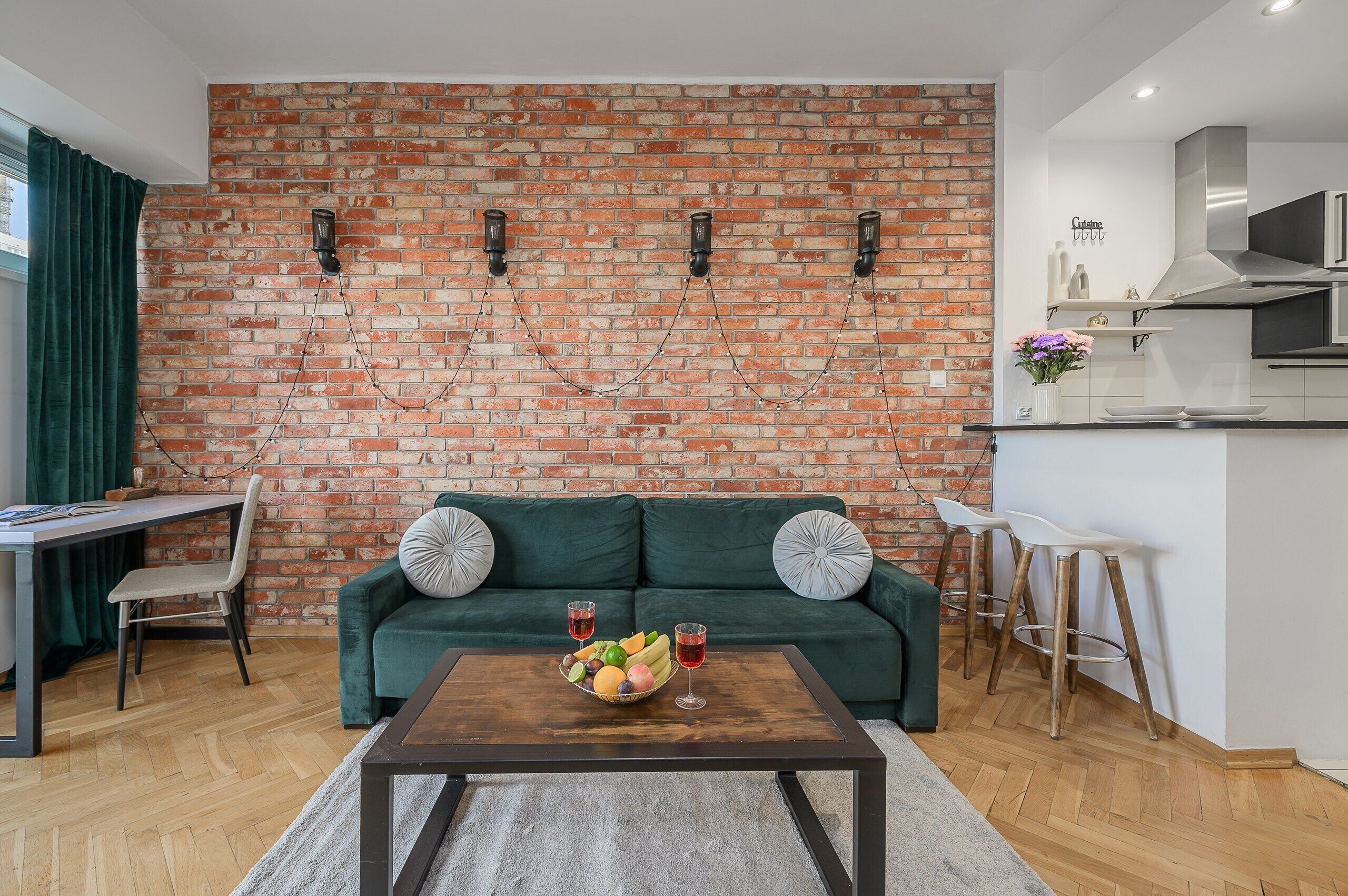 Uma sala de estar moderna com parede de tijolos, sofá verde, mesa de centro de madeira com frutas, e área de cozinha com armários brancos e coifa de aço inoxidável.
