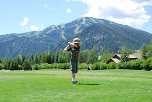 Uma pessoa vestida com roupas de camuflagem segurando um taco de golfe em um campo gramado, com uma montanha ao fundo.