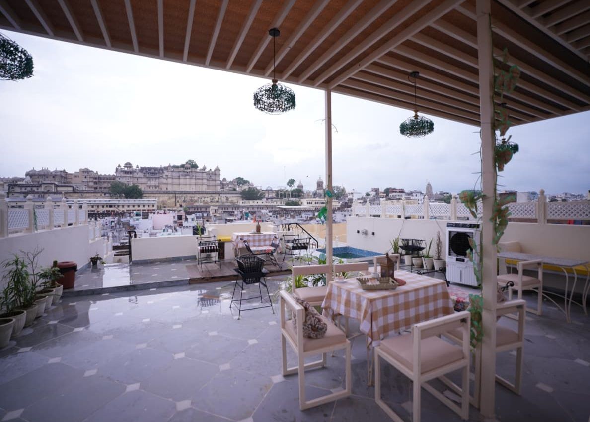 Terraço no telhado com mesas, cadeiras, plantas em vasos e vista para a cidade.