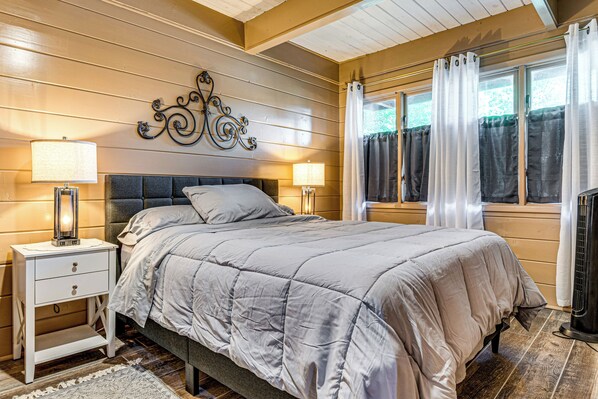 Quarto com cama, duas mesas de cabeceira com luminárias, um objeto decorativo na parede e uma janela com cortinas.