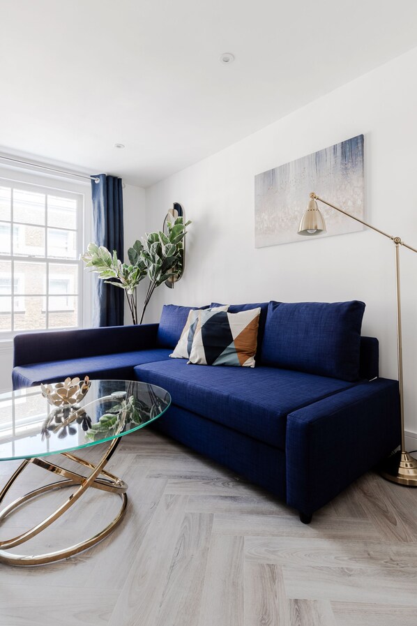Uma sala de estar moderna com um sofá azul, uma mesa de centro de vidro com pés dourados, um abajur e uma grande janela com cortinas.