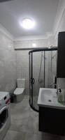 Casa de banho com cabine de duche em vidro, sanita, lavatório e máquina de lavar roupa.