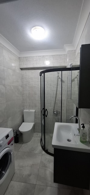 Casa de banho com cabine de duche em vidro, sanita, lavatório e máquina de lavar roupa.