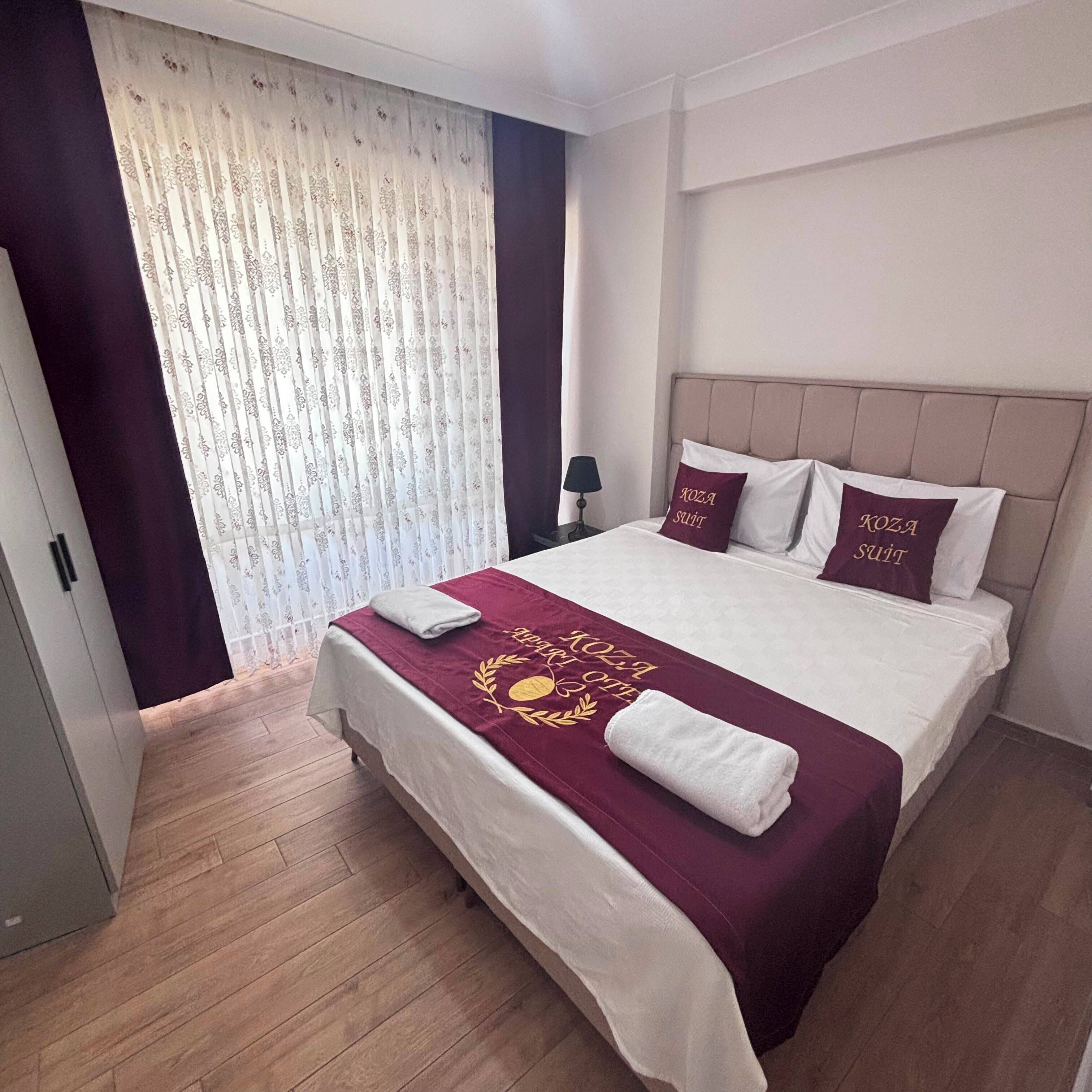 Quarto de hotel com cama, mesa de cabeceira, abajur e janela com cortinas.