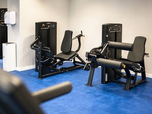 Uma academia com equipamentos de exercício, um dispensador de álcool em gel fixado na parede e uma máquina branca.