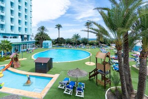 Área de piscina em resort, com piscina grande, espreguiçadeiras, palmeiras e um prédio ao fundo.