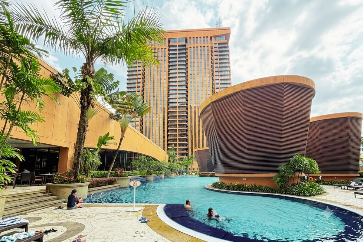 Uma área de piscina com um edifício curvo, palmeiras e um hotel alto ao fundo.