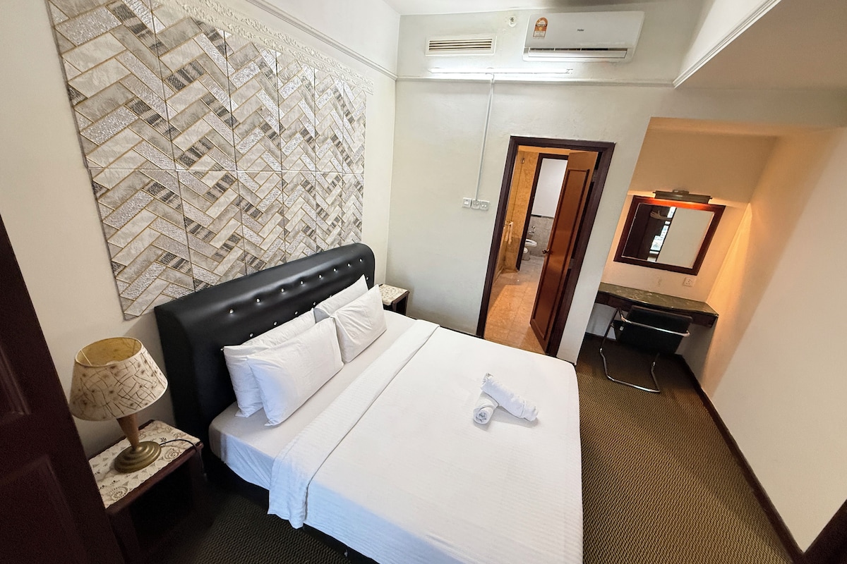 Quarto de hotel com cama, mesa de cabeceira, luminária, espelho e banheiro visível por uma porta aberta.