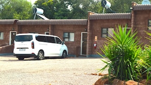 Uma carrinha branca estacionada em frente a um edifício com entrada de automóveis em cascalho, uma antena parabólica e um extintor fixado na parede.