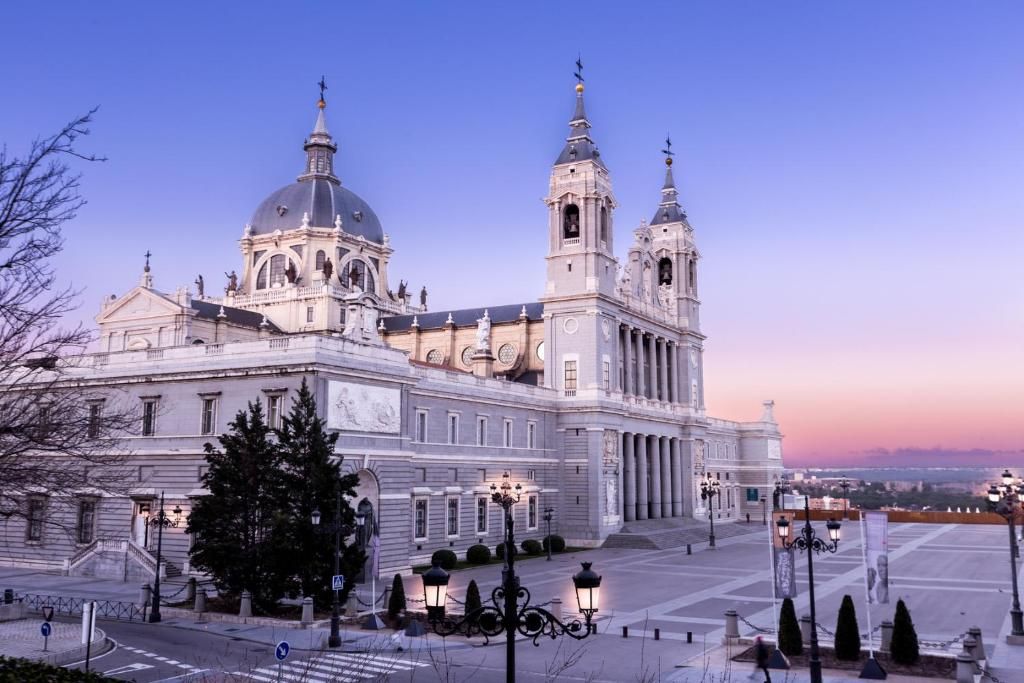 Um edifício grande e histórico com uma cúpula imponente e duas torres, rodeado por uma ampla praça com postes de luz e algumas árvores.
