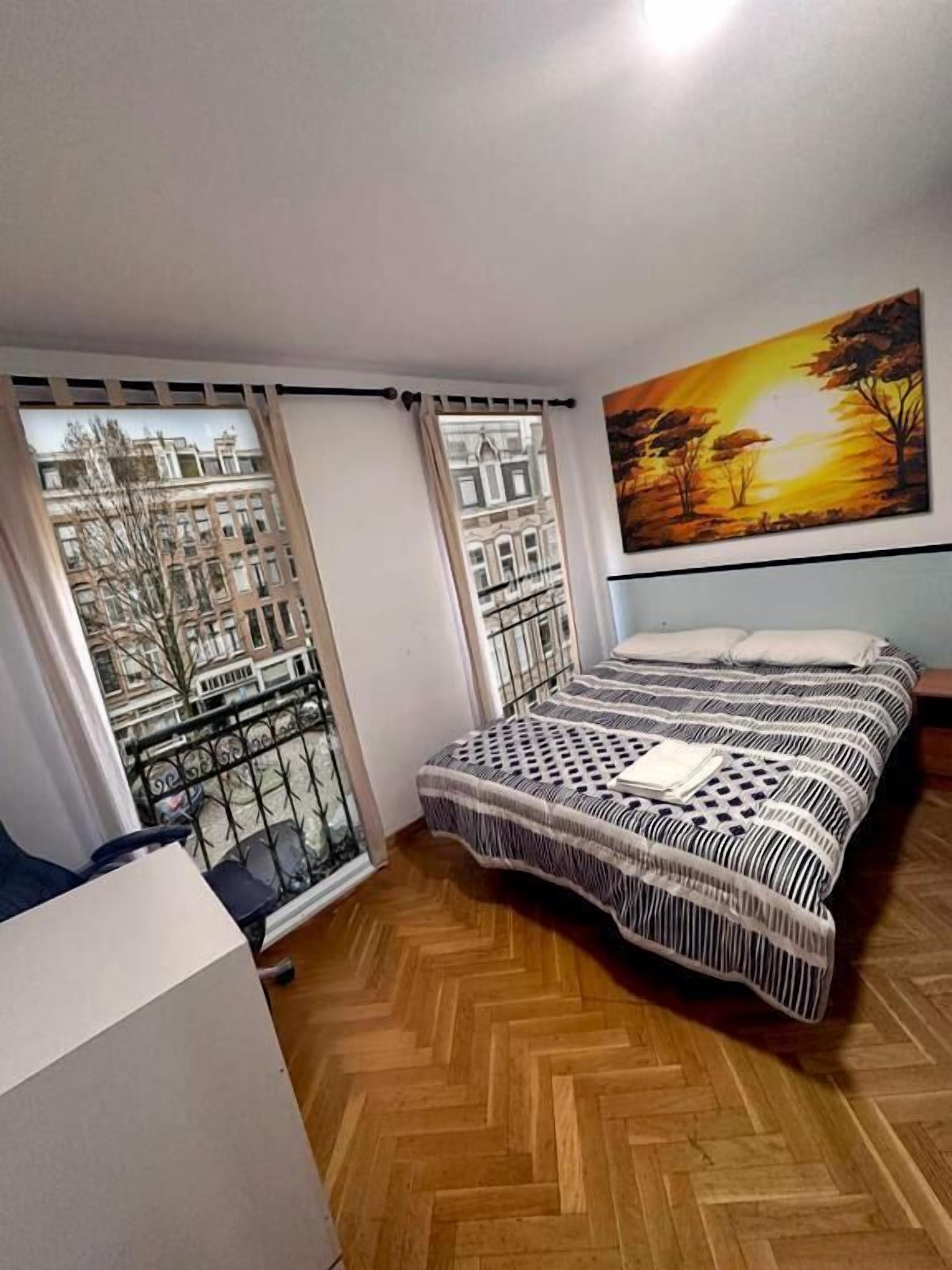 Quarto com cama, janela com cortinas, vista de prédios e um quadro na parede.