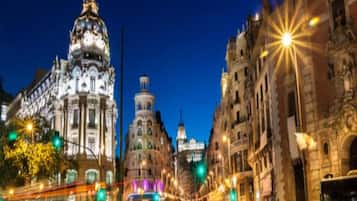 Paisagem urbana noturna com edifícios históricos, uma estrutura abobadada em destaque e uma rua movimentada com semáforos.