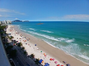 Uma praia com guarda-sóis e pessoas, céu claro e edifícios ao fundo.