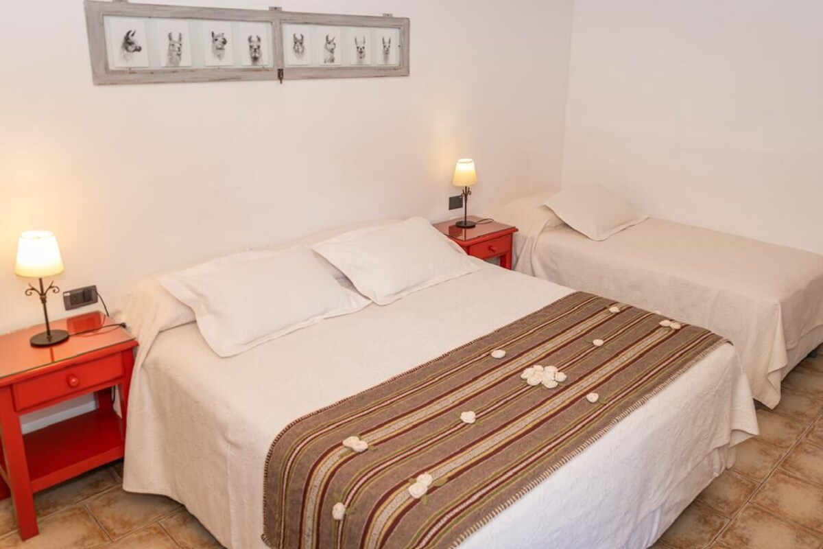 Quarto de hotel com duas camas, uma mesa de cabeceira vermelha com uma lâmpada, um quadro na parede e um espelho.