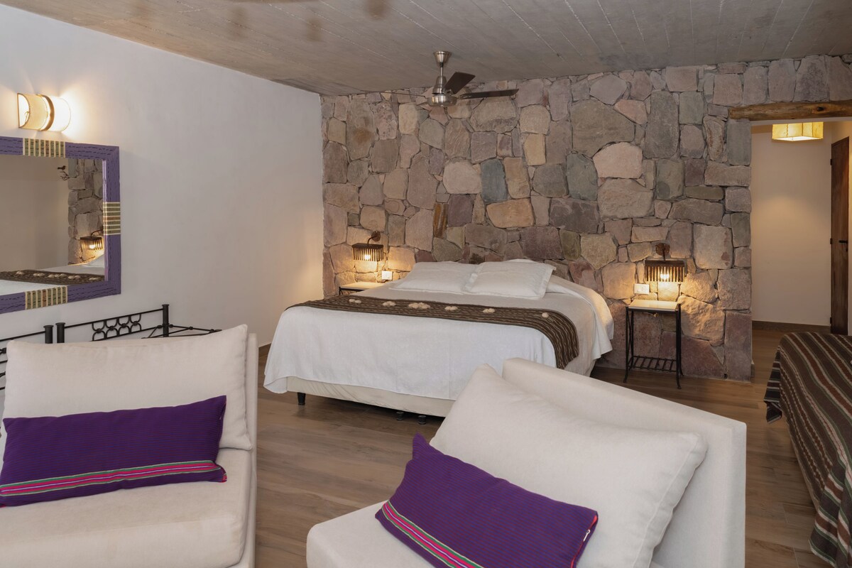 Quarto com parede de pedra, duas camas, ventilador de teto e mesa de cabeceira com abajur.