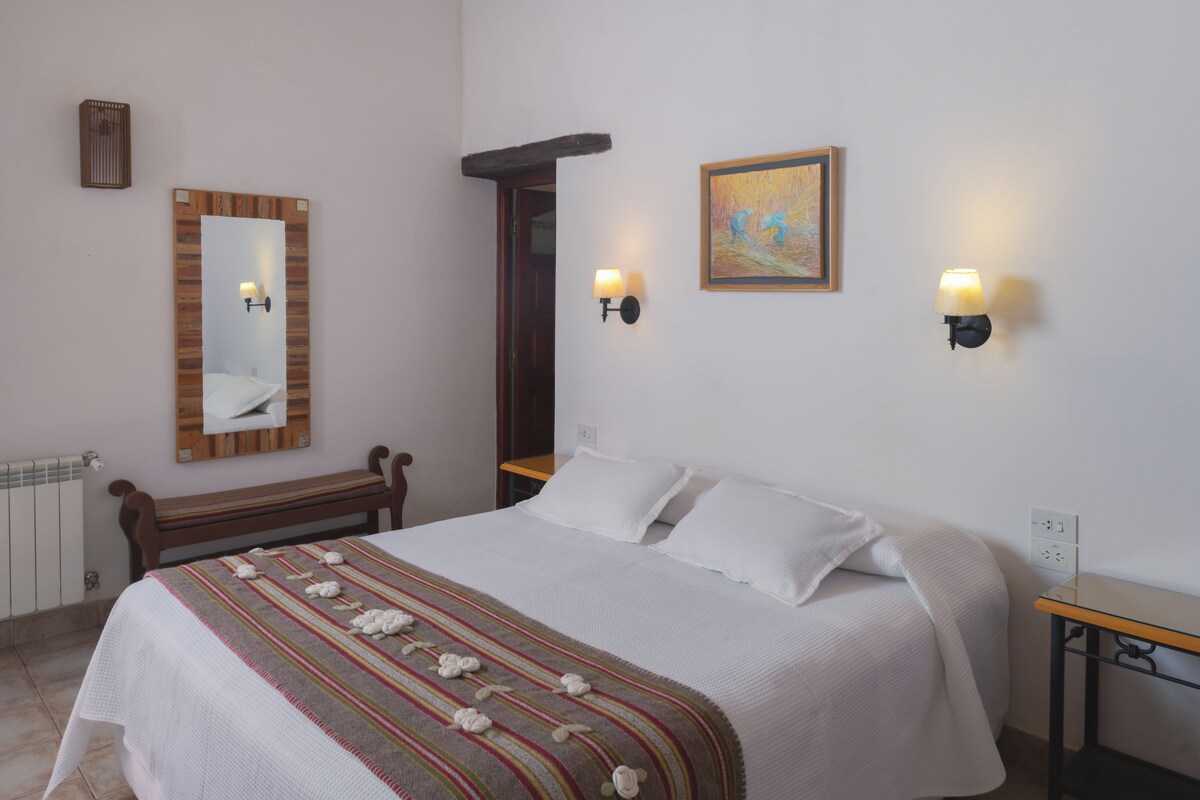 Quarto de hotel com uma cama, um banco, um espelho, luminárias na parede e um quadro.