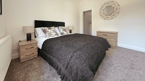 Quarto com cama, mesas de cabeceira, cômoda, espelho e decoração geométrica na parede.