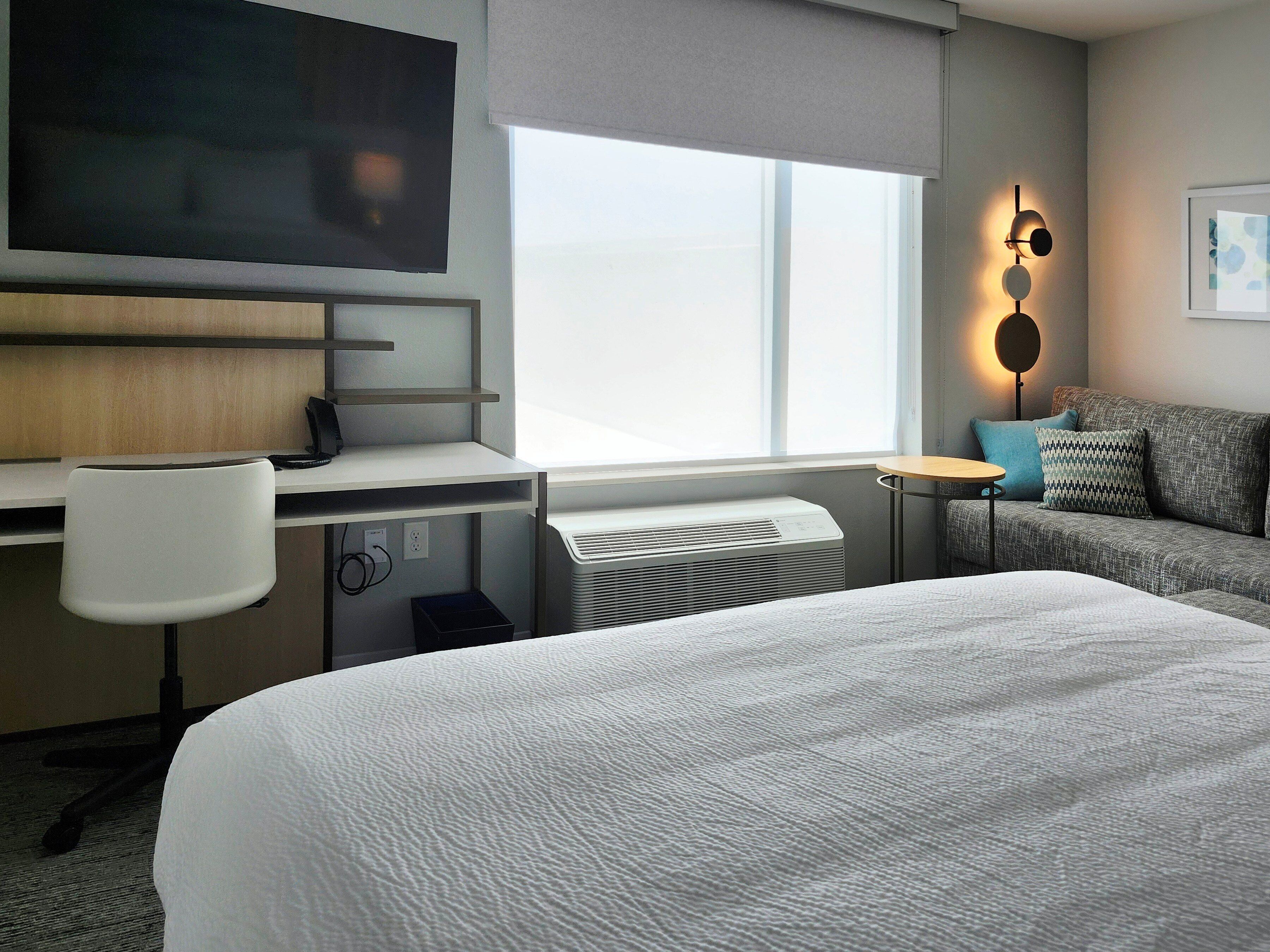 Quarto de hotel com cama, escrivaninha com cadeira, televisão, aquecedor, sofá com almofadas e janela com persianas.