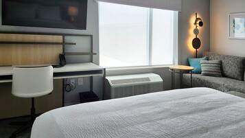 Quarto de hotel com cama, escrivaninha com cadeira, televisão, aquecedor, sofá com almofadas e janela com persianas.