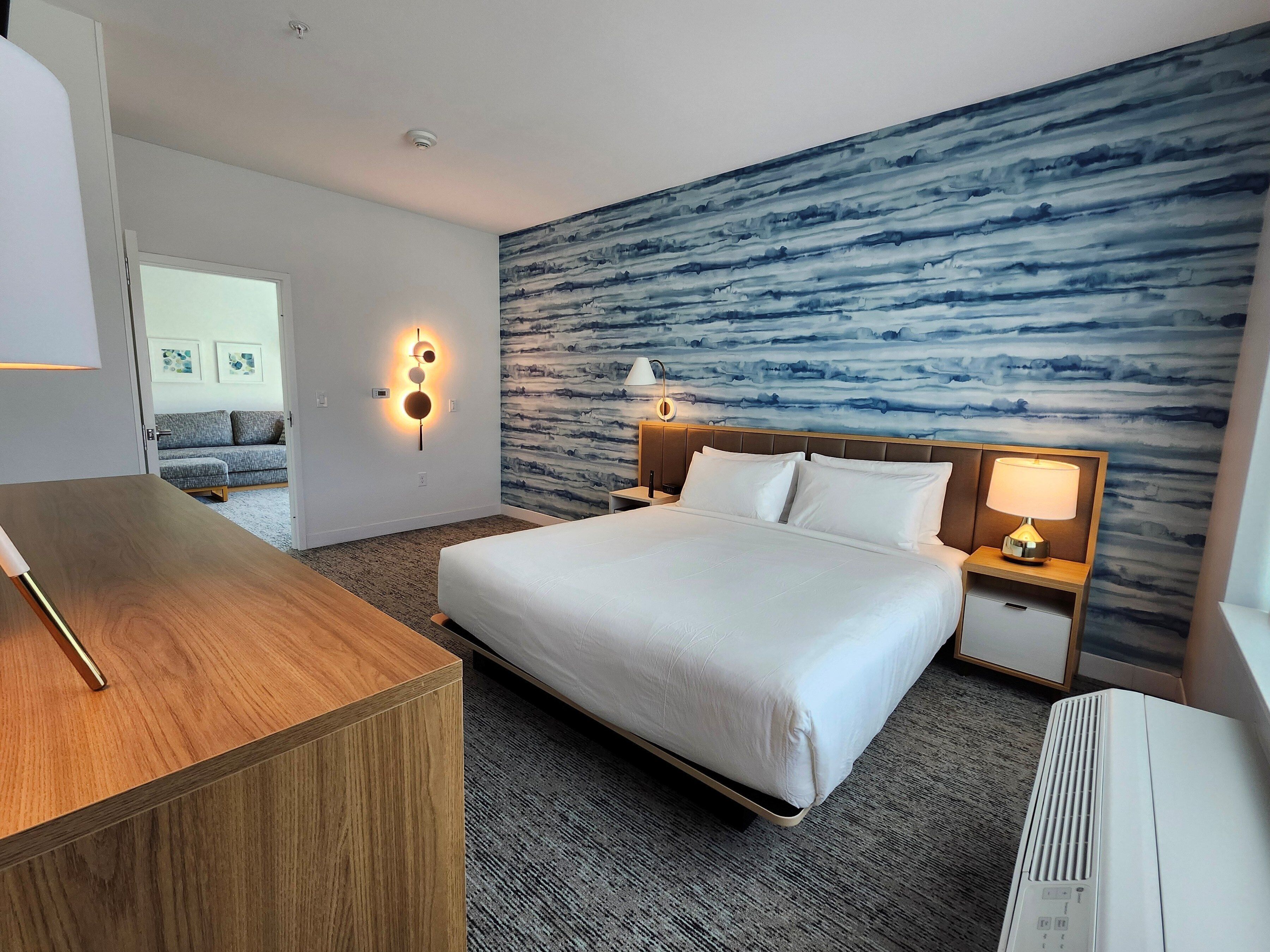 Quarto de hotel com cama, mesas de cabeceira, escrivaninha e parede com design abstrato azul.