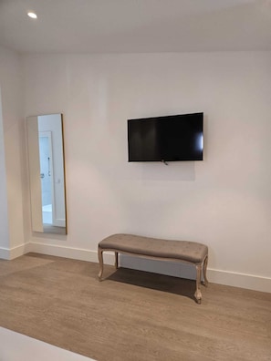 Quarto com televisão de tela plana presa na parede, um banco e uma porta que dá para outro cômodo.