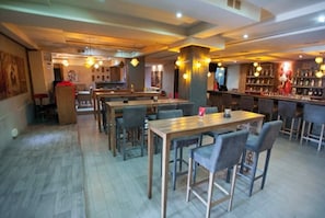 Interior de restaurante com várias mesas e cadeiras para refeições, área de bar com prateleiras cheias de garrafas e iluminação pendente.