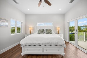 Quarto com uma cama grande, duas mesas de cabeceira com luminárias, um ventilador de teto e vista através de uma porta de vidro deslizante.