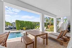 Uma área de piscina moderna ao ar livre com mesa de madeira e cadeiras de descanso, cercada por um muro branco e vista para uma paisagem tropical.
