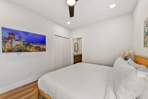 Quarto com uma cama grande, uma mesinha de cabeceira, um espelho e uma pintura de uma paisagem urbana.