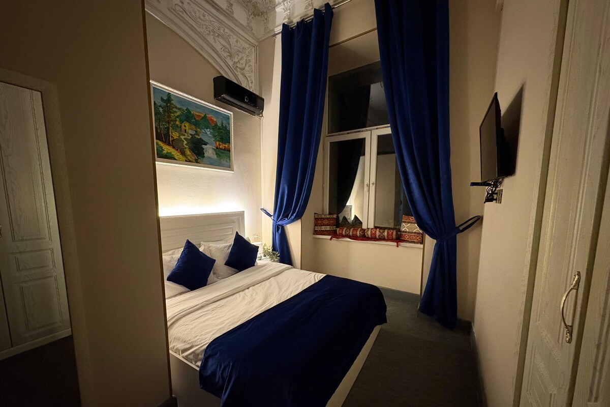 Um quarto com cama, um quadro, uma janela com cortinas e uma TV na parede.