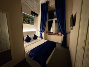 Um quarto com cama, um quadro, uma janela com cortinas e uma TV na parede.