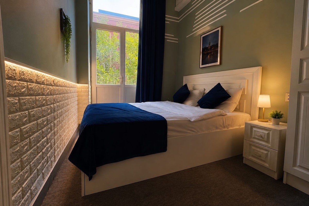 Quarto com cama grande, mesa de cabeceira com abajur, quadro na parede e janela com cortinas.