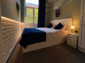 Quarto com cama grande, mesa de cabeceira com abajur, quadro na parede e janela com cortinas.