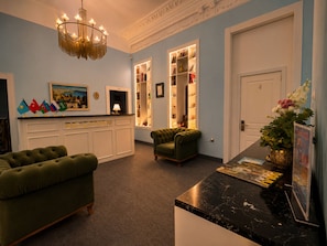 Uma área de recepção com um candelabro, um balcão de atendimento, uma poltrona verde e uma estante repleta de livros e objetos decorativos.