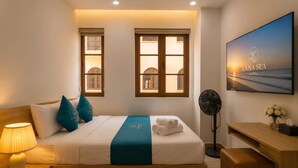 Quarto de hotel com cama, escrivaninha, cadeira, ventilador e televisão mostrando uma cena de praia.