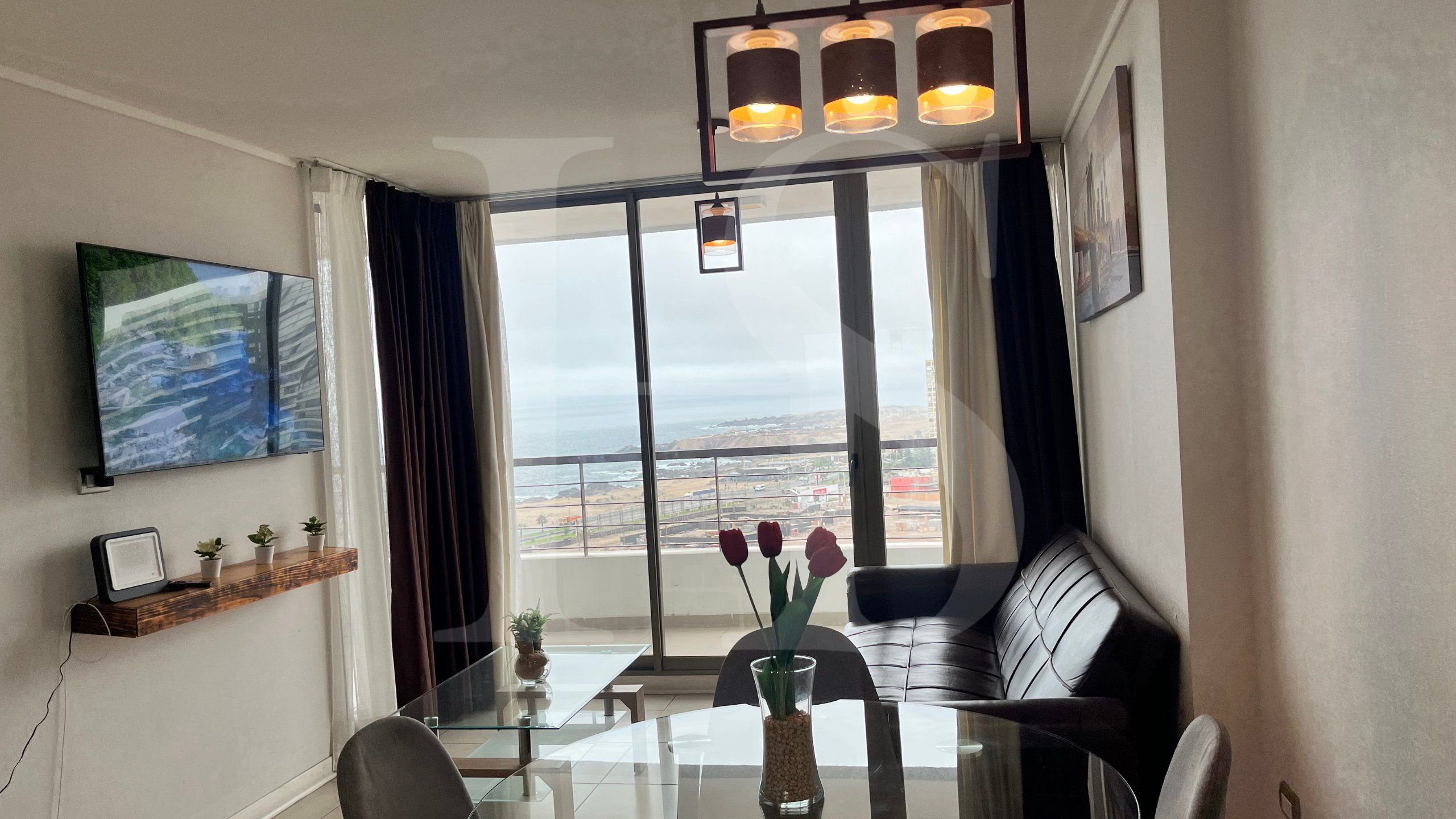 Uma sala de estar moderna com mesa de vidro, sofá de couro preto, TV montada na parede e vista para a paisagem urbana por grandes janelas.