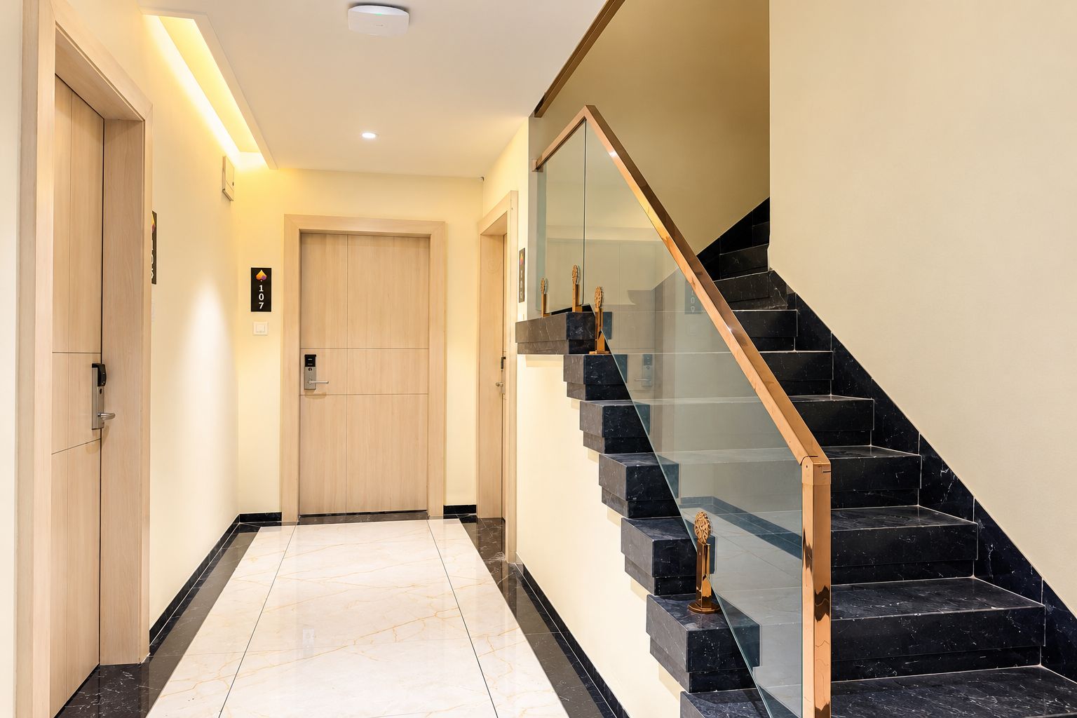 Um corredor moderno com porta de madeira, escada com guarda-corpo de vidro e chão de mármore.