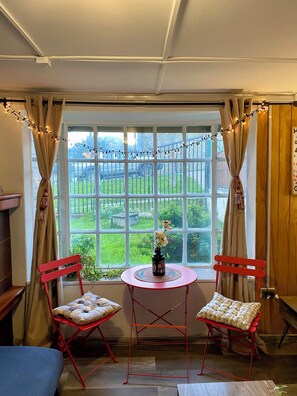 Quarto com duas cadeiras vermelhas, uma mesa redonda pequena com um vaso, e vista para uma paisagem verde através de uma grande janela.