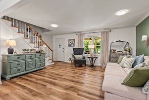Uma sala de estar ampla com piso de madeira, uma escada, um aparador verde, uma poltrona cinza e um sofá bege.