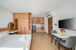 Quarto de hotel com cama, escrivaninha, cadeira, televisão, espelho e cozinha compacta.