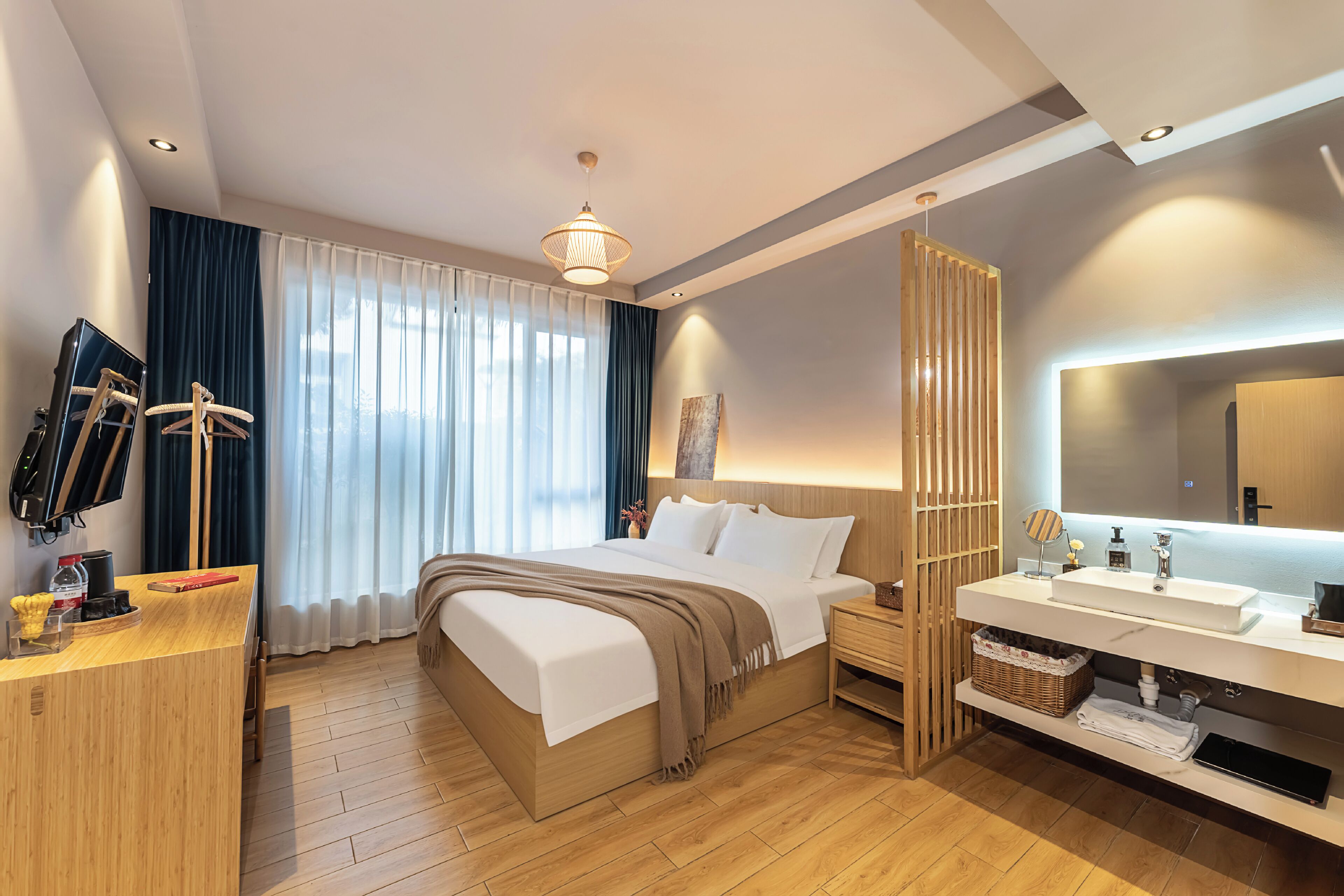 Quarto de hotel moderno com uma cama grande, uma mesinha de cabeceira, uma escrivaninha com televisão de tela plana, um guarda-roupa de madeira e banheiro com um espelho grande e pia.