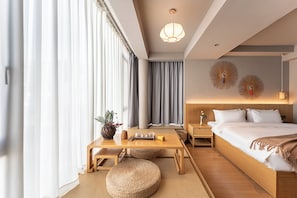 Quarto de hotel moderno com uma cama grande, mesas de cabeceira, uma escrivaninha de madeira, um banquinho tecido e vista para o exterior através de grandes janelas.