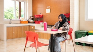 Mulher sentada à mesa com um recipiente rosa, num quarto com armários de madeira e uma janela.