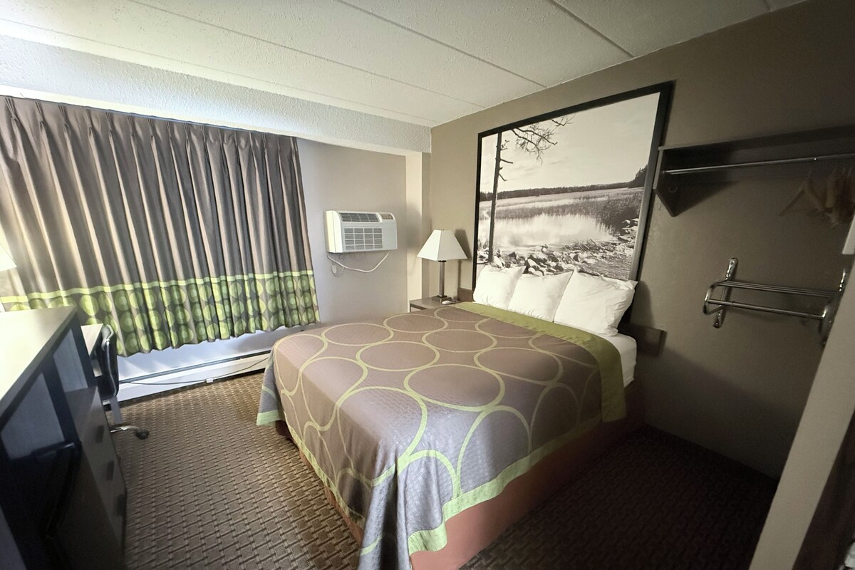 Quarto de hotel com cama, escrivaninha, cadeira, janela com cortinas, ar-condicionado, luminária e um quadro na parede.