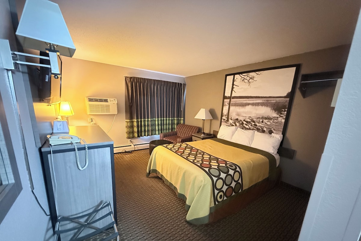 Quarto de hotel com cama, escrivaninha com telefone, luminária, janela com cortinas e um quadro na parede.