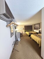 Quarto de hotel com cama, escrivaninha, cadeira, cabideiro e janela com cortinas.