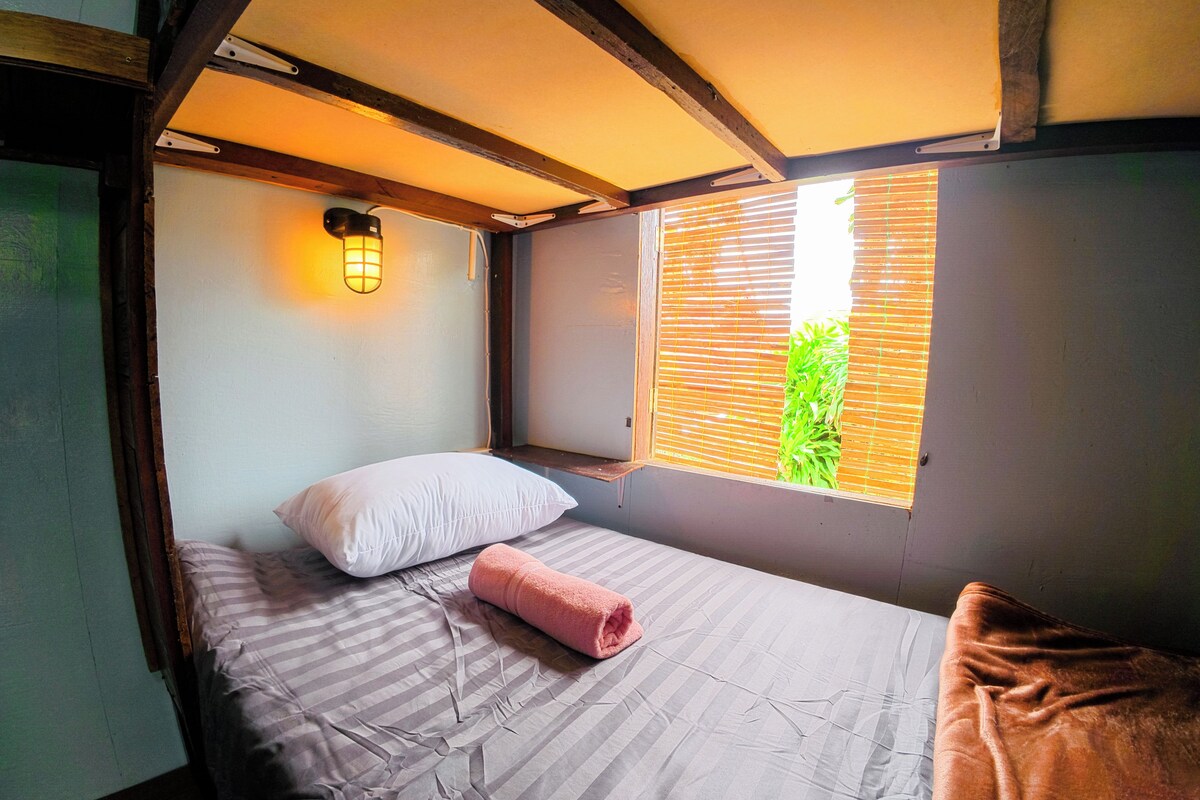 Uma cama de solteiro com colchão listrado, um travesseiro branco e uma toalha rosa. Há uma janela com persianas, um aparelho de luz fixado na parede e um vigamento de madeira no teto.