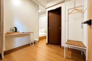 Quarto de hotel com piso de madeira, uma cama, um guarda-roupa, uma mesinha com chaleira e um cabideiro.